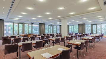 Uma sala de conferências com várias mesas e cadeiras, janelas amplas e piso coberto por um tapete vermelho.