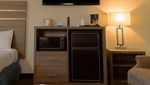 Quarto de hotel com cama, televisão na parede, micro-ondas, mini geladeira, luminária e pequena mesa com cafeteira.