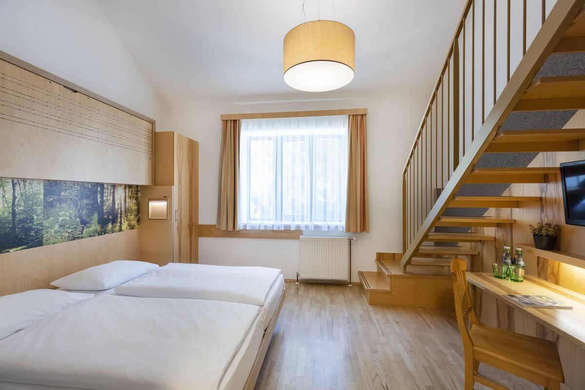 Quarto de hotel com duas camas, uma janela grande com cortinas, uma escada de madeira e uma televisão presa à parede.