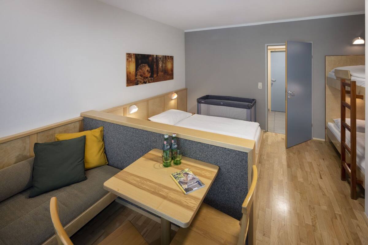 Quarto compacto com beliche, um pequeno sofá, uma mesa com bebidas e um quadro na parede.