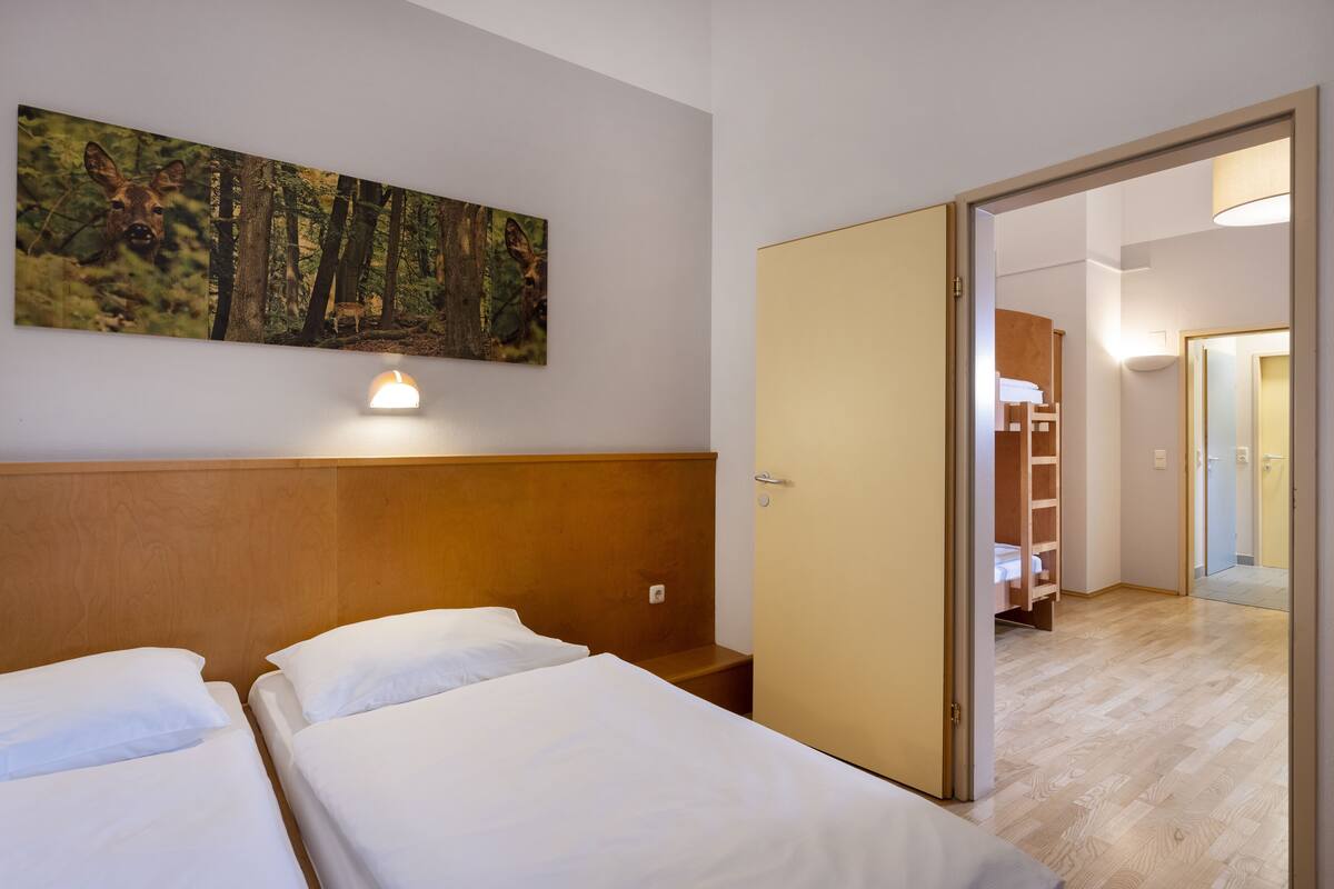 Quarto de hotel com duas camas, cabeceira de madeira, pintura de uma floresta e corredor que dá para um banheiro.