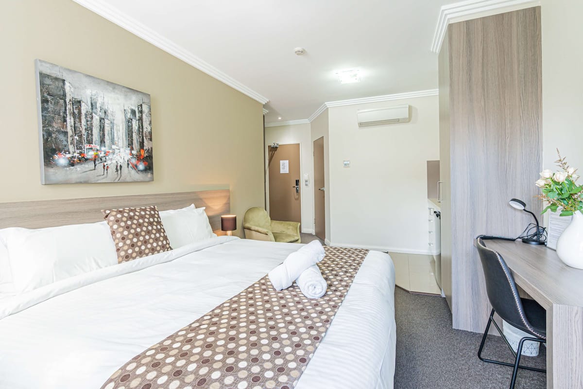 Quarto de hotel com uma cama grande, uma escrivaninha, uma cadeira, um quadro na parede e uma porta que dá para outro cômodo.