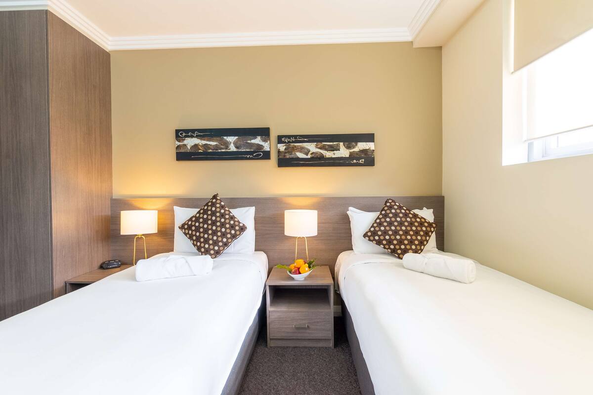 Quarto de hotel com duas camas, uma mesa de cabeceira com abajur, uma pequena mesa com frutas e uma obra de arte na parede.