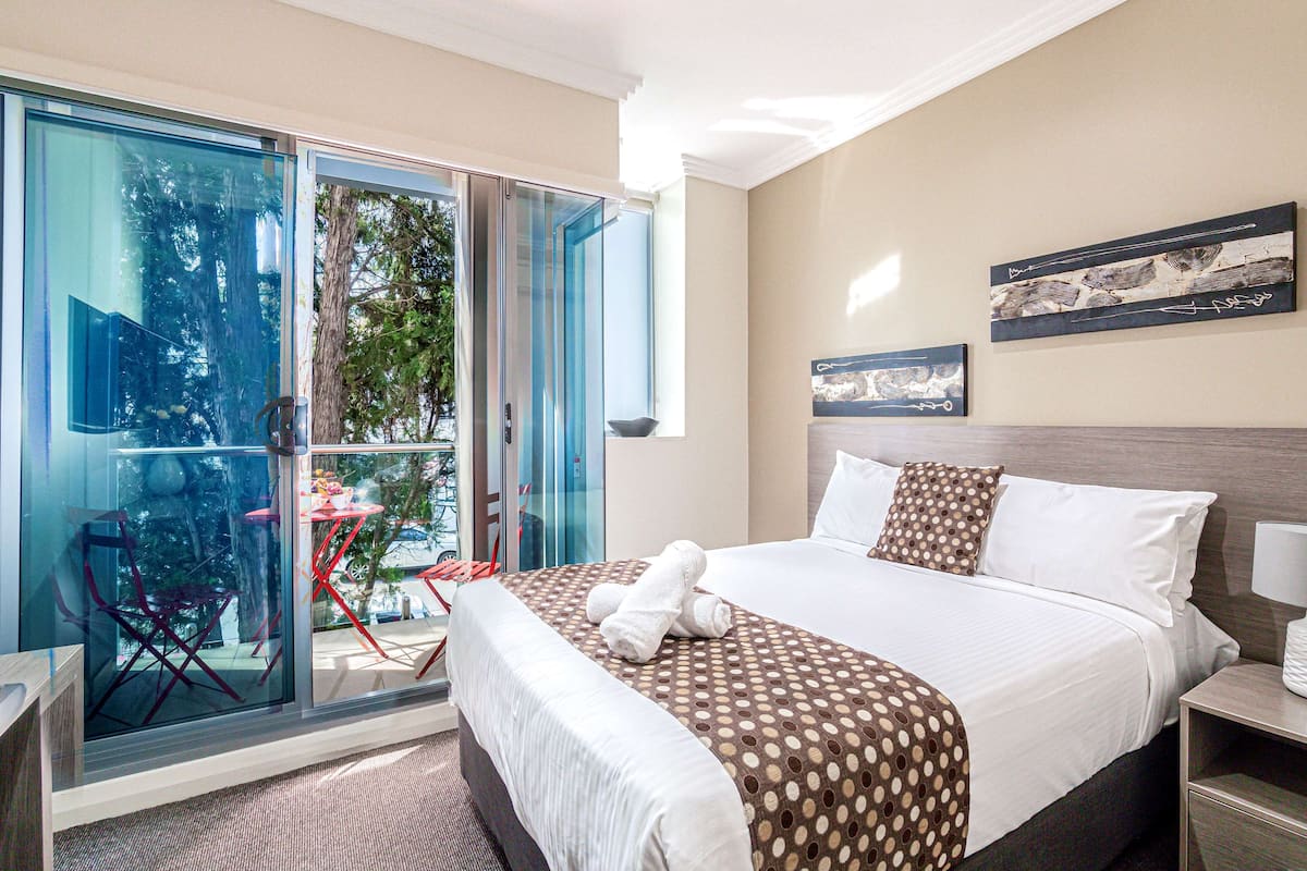 Quarto de hotel com uma cama grande, uma mesa de cabeceira, uma cadeira e uma porta de vidro deslizante para uma área externa com mesa e cadeiras.