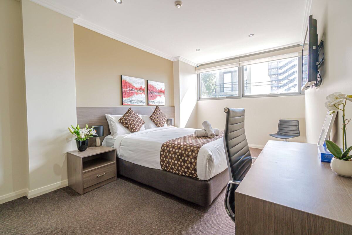 Quarto de hotel com cama, escrivaninha, cadeira, televisão e janela.
