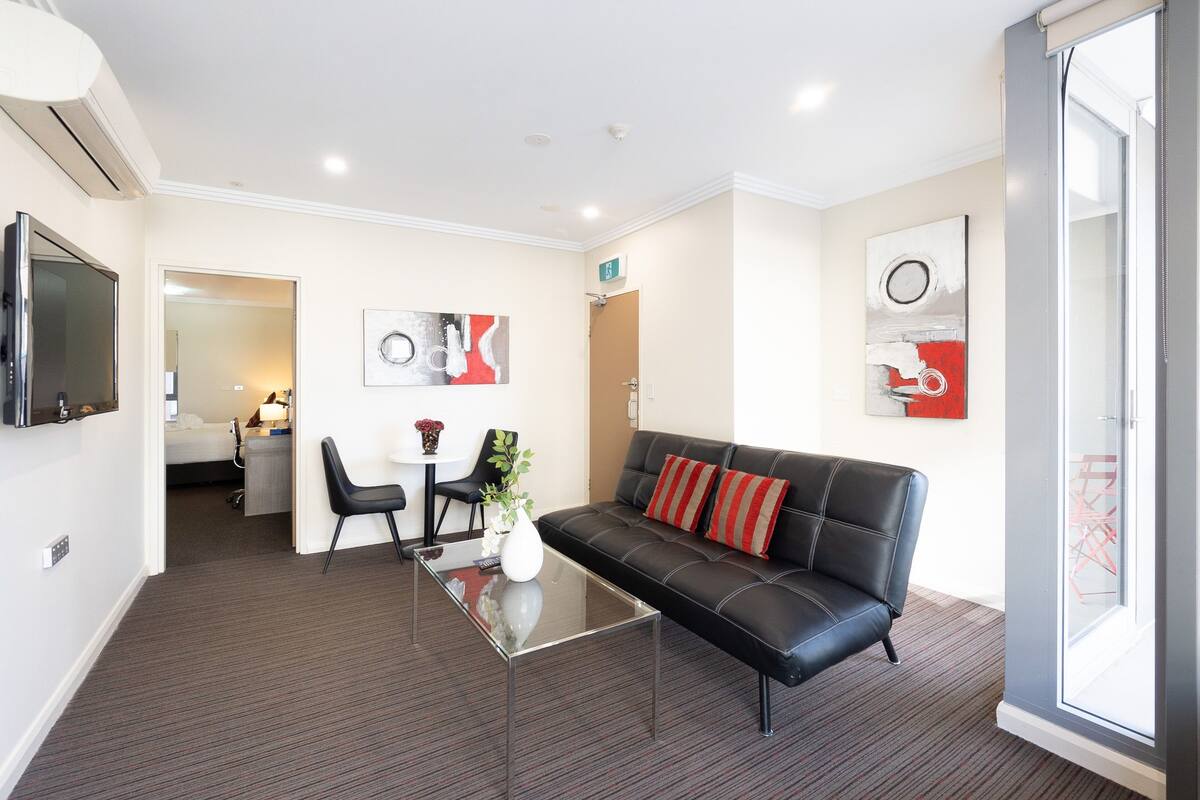 Quarto de hotel moderno com um sofá de couro preto, uma mesa de centro de vidro e uma televisão de tela plana. Há uma área de jantar com mesa e cadeiras, e uma pequena planta na mesa de centro.
