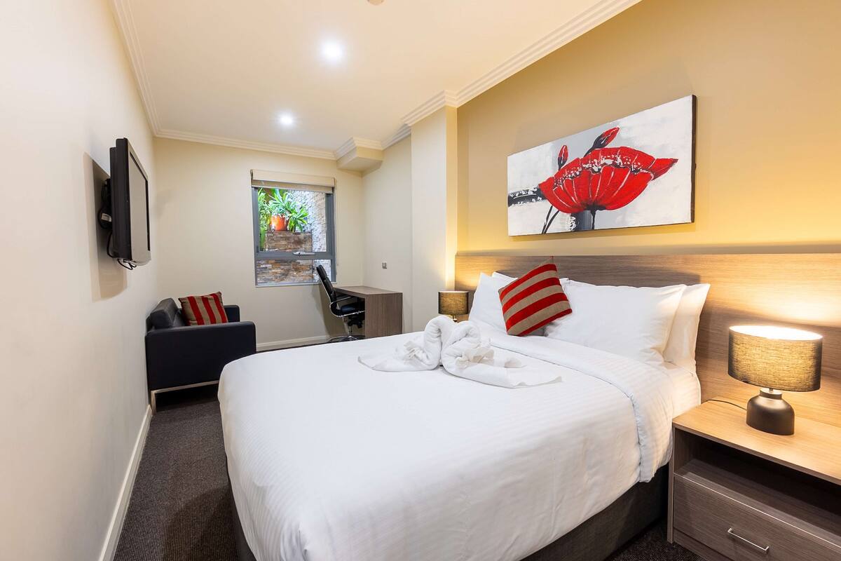 Quarto de hotel com uma cama grande, uma mesa, uma cadeira, televisão, janela com vista para o verde e um quadro na parede.