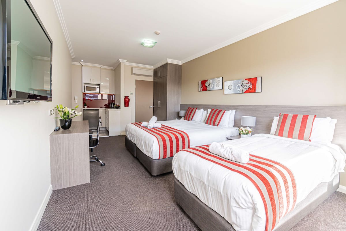 Quarto de hotel com duas camas, uma mesa, televisão e cozinha compacta.