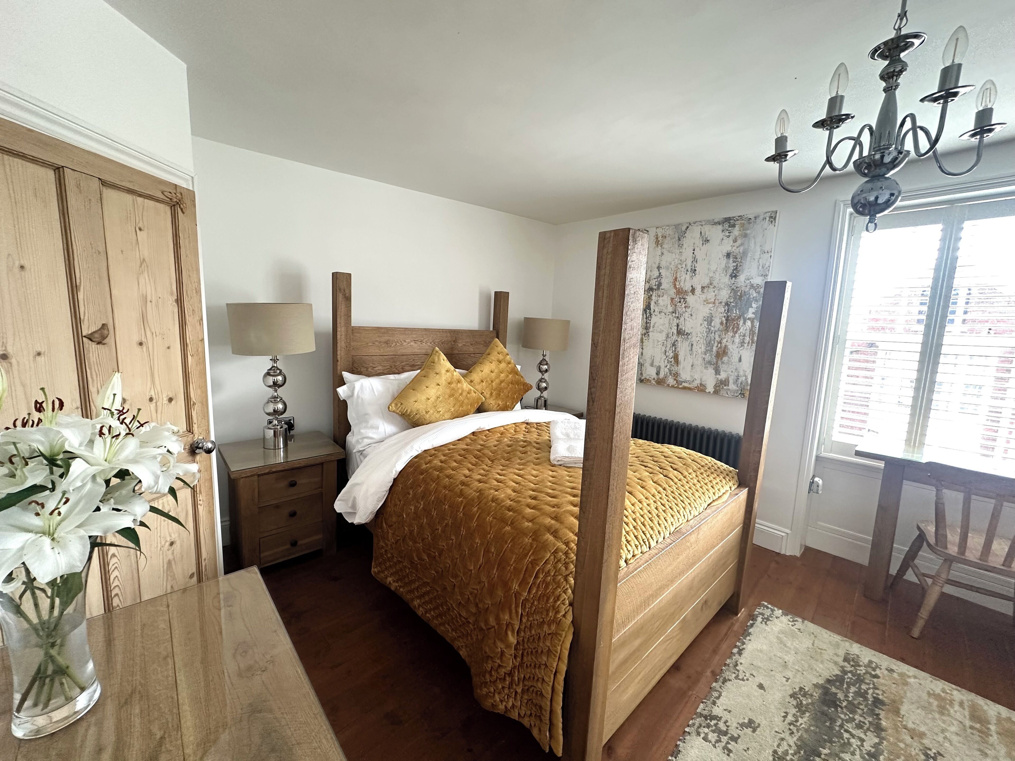Quarto com cama de madeira, roupa de cama branca e dourada, mesa de cabeceira com abajur, um vaso de flores e vista para o exterior pela janela.