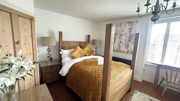 Quarto com cama de madeira, roupa de cama branca e dourada, mesa de cabeceira com abajur, um vaso de flores e vista para o exterior pela janela.