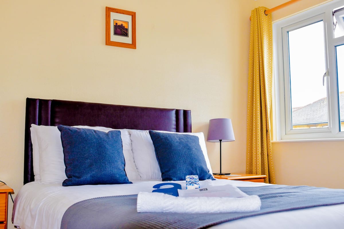 Uma cama bem arrumada com almofadas azuis e brancas, uma mesa de cabeceira com abajur e uma janela com cortinas amarelas.