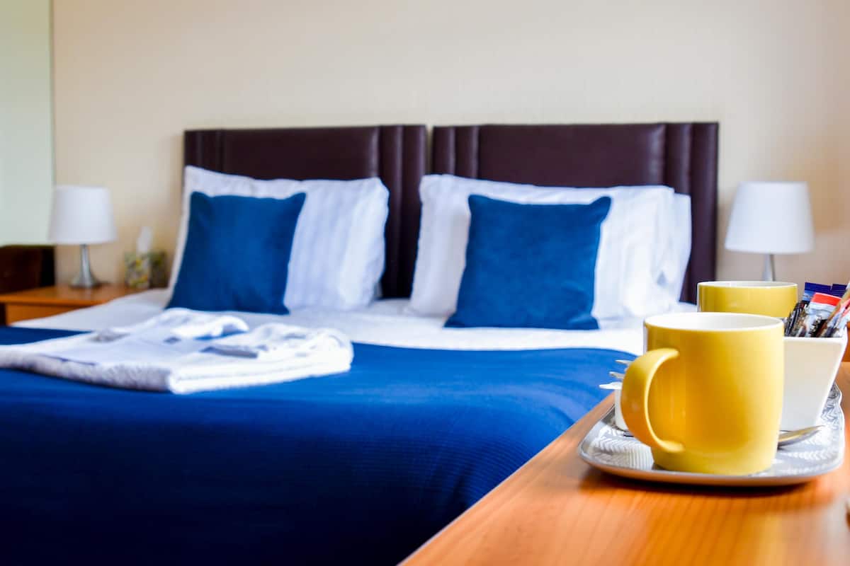 Quarto de hotel com uma cama, dois travesseiros, um cobre-leito azul, uma caneca amarela sobre uma bandeja e uma toalha branca.