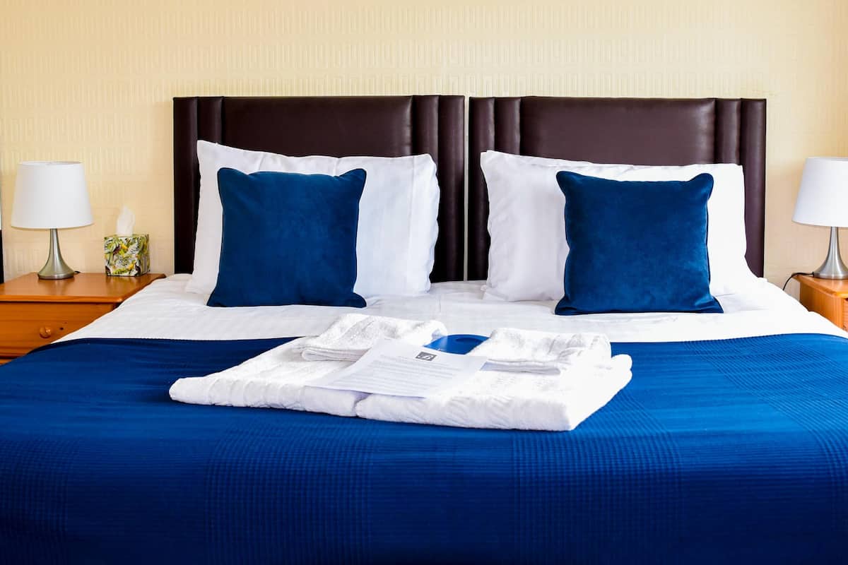 Cama bem arrumada com colcha azul, lençóis brancos e duas almofadas azuis. Dois mesinhas de cabeceira, com luminárias e uma caixa de lenços em uma delas.