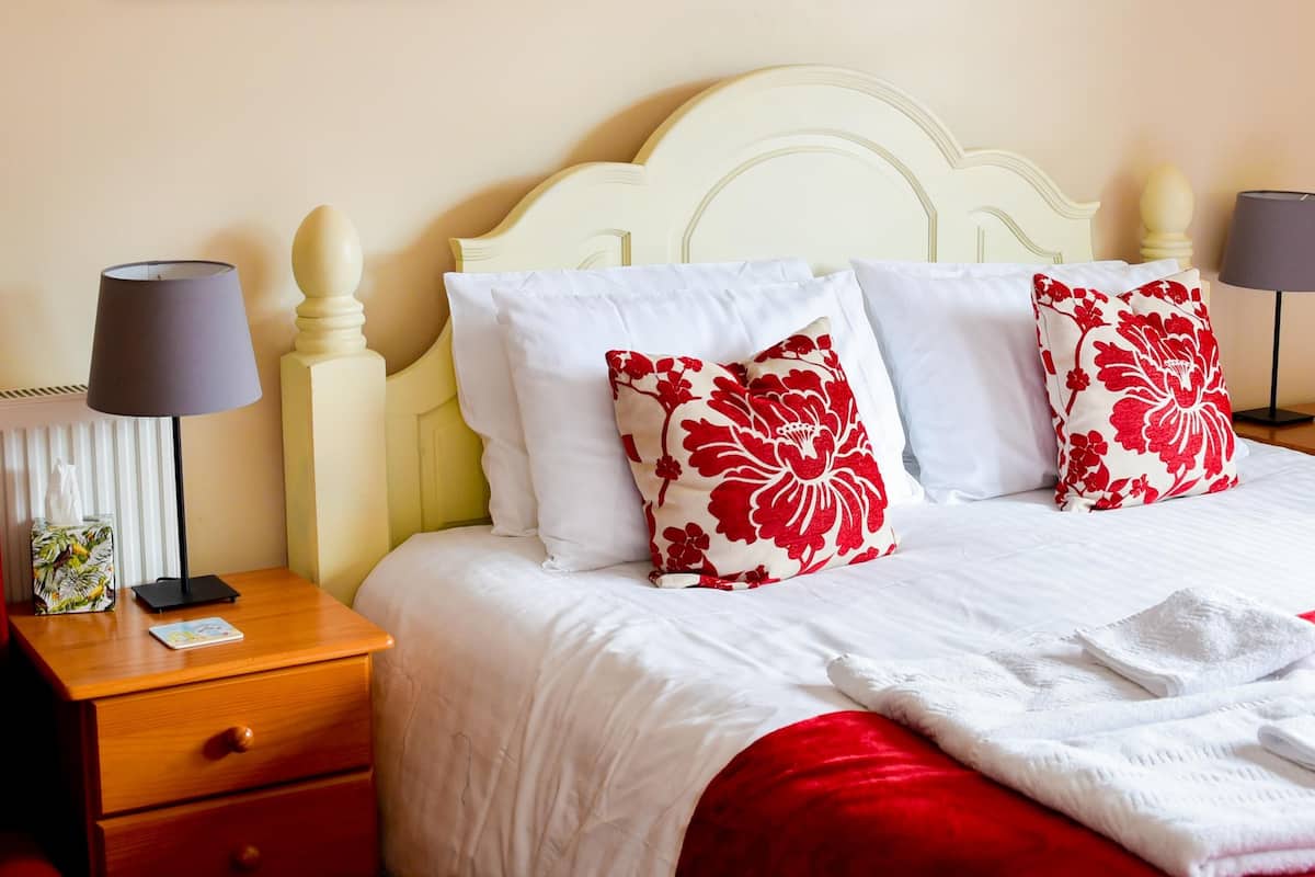 Cama bem arrumada com almofadas florais brancas e vermelhas, um cobertor vermelho e uma cabeceira branca. Há duas mesas de cabeceira com abajures e um pequeno criado-mudo de madeira com um estojo de lenços e um cartão.
