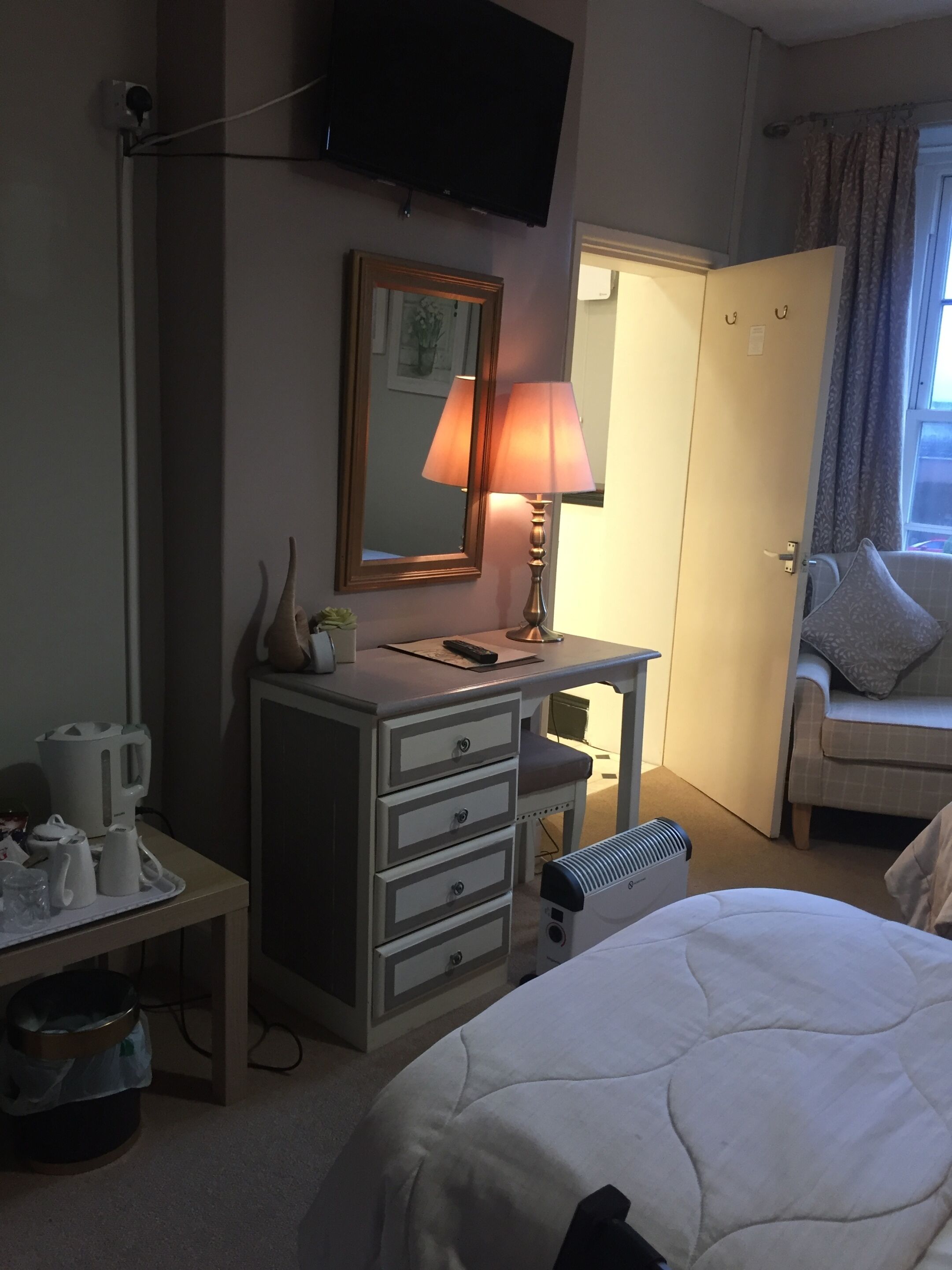 Quarto de hotel com uma cama, uma escrivaninha com candeeiro, televisão, guarda-roupa e janela com cortinas.