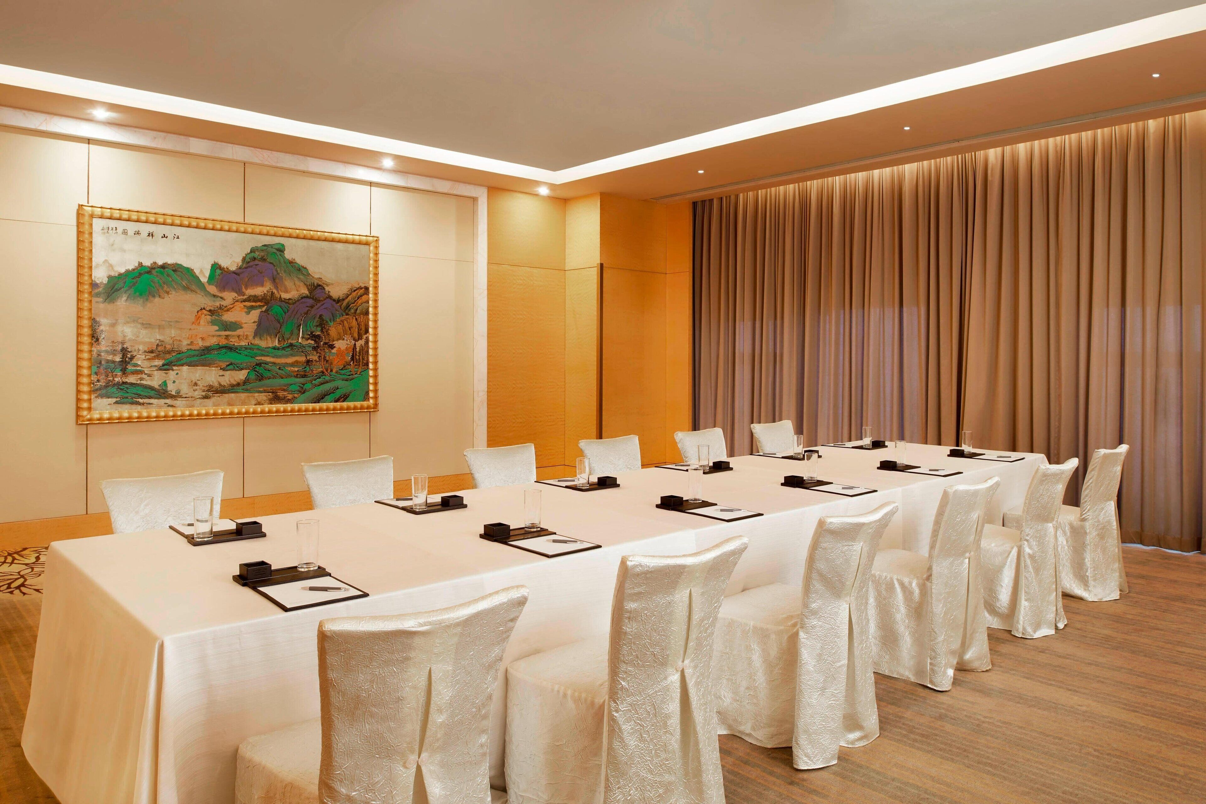 Uma sala de conferência com uma mesa comprida, cadeiras brancas e um quadro grande emoldurado na parede.