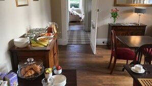 Uma área de refeições com buffet de café da manhã, uma mesa de jantar com abajur e vista para o jardim através de uma porta aberta.
