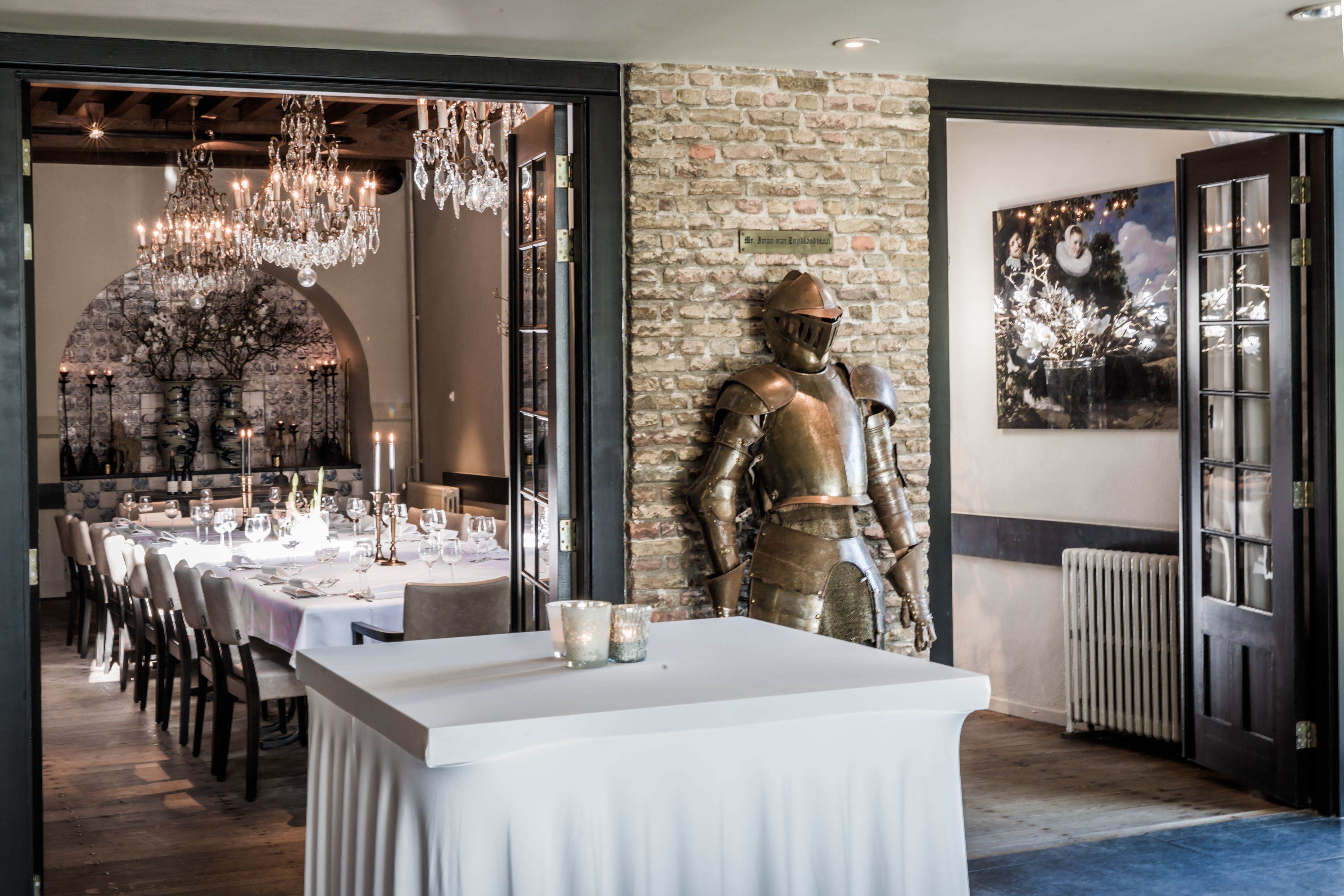 Uma sala de jantar com uma mesa comprida posta para refeição, um candelabro e uma estátua de cavaleiro.