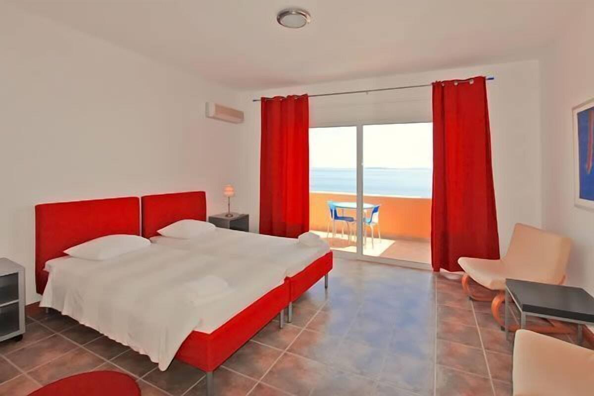 Quarto de hotel com cama, uma pequena mesa, uma cadeira e vista para o mar através de uma varanda.