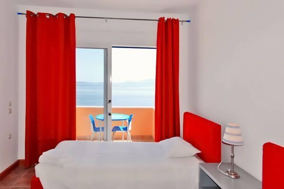 Quarto de hotel com cama, mesa e cadeiras, luminária e vista para o mar.