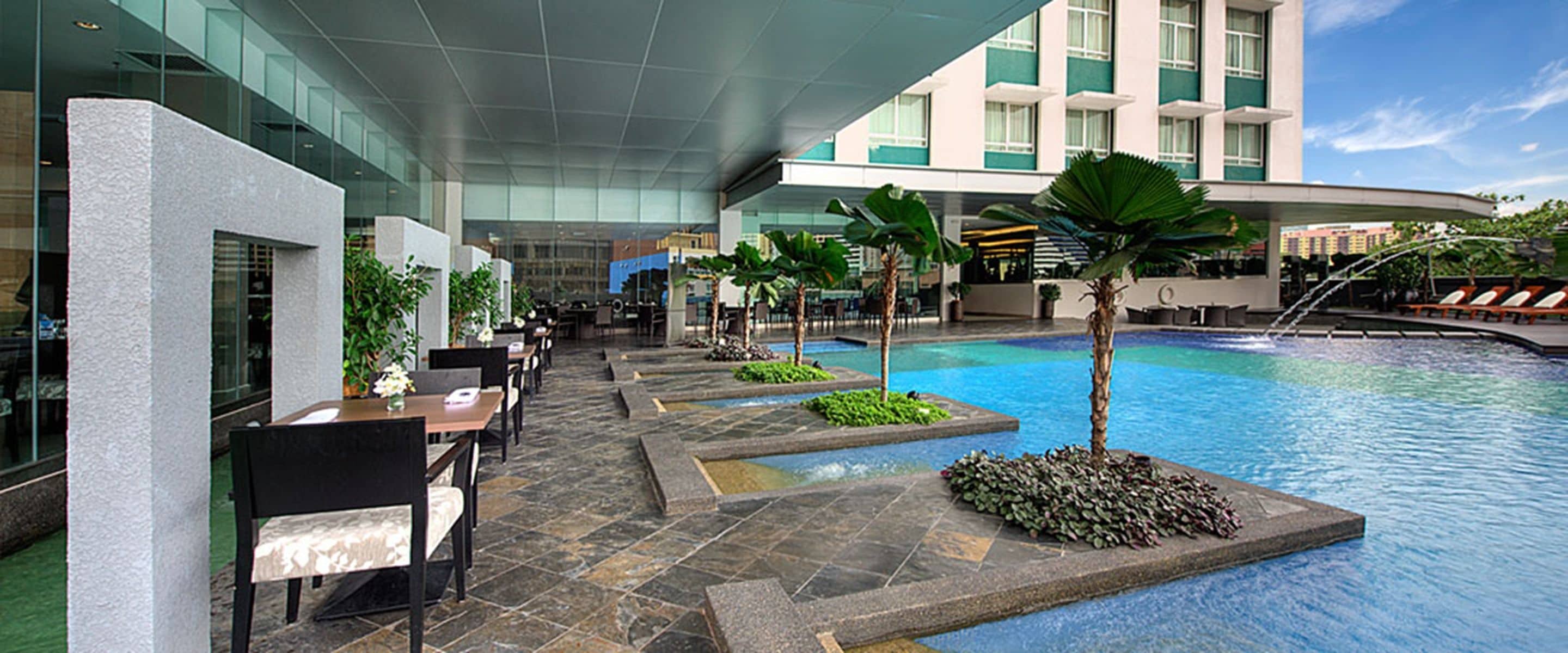 Área de piscina de hotel com espaço para sentar ao ar livre e um edifício moderno ao fundo.