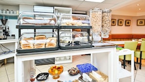 Um buffet de café da manhã com diversos alimentos, incluindo pão, doces e frutas, em uma sala de jantar bem iluminada e com assentos.