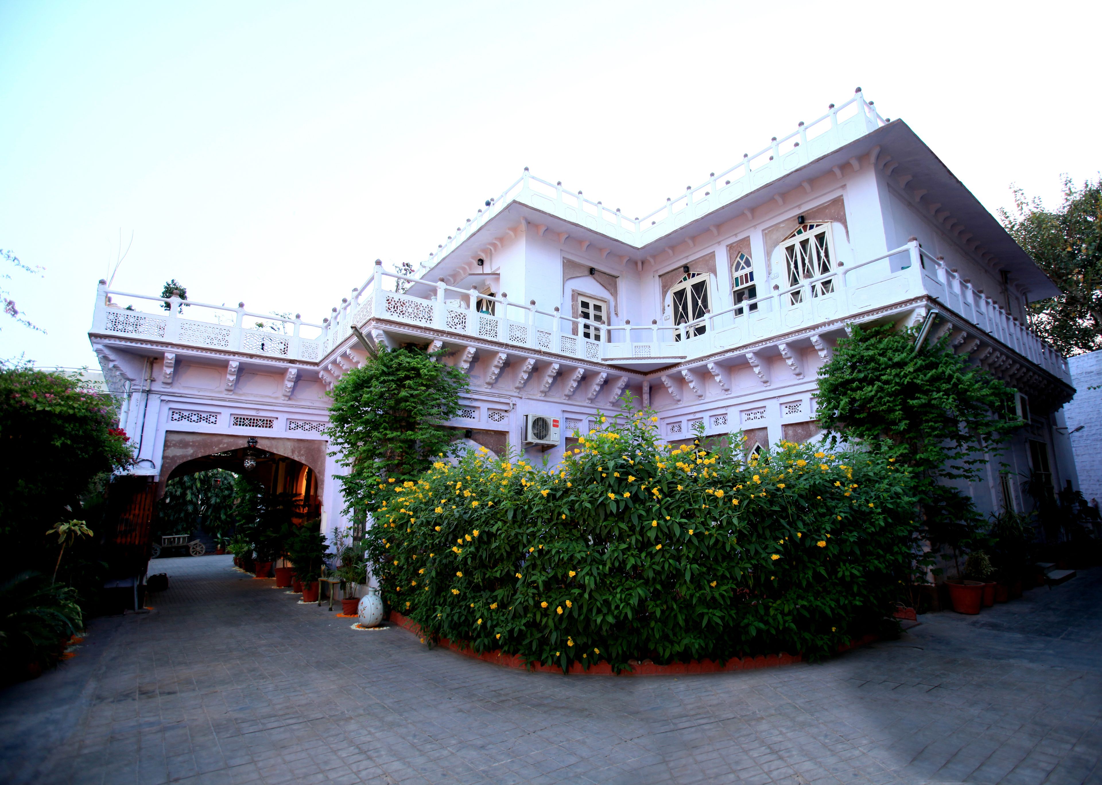Um edifício tradicional com um arco central, uma varanda com grades elaboradas e um jardim com flores amarelas.
