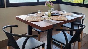 Uma mesa de jantar com quatro cadeiras, um vaso com flores e vista para o mar através de grandes janelas.