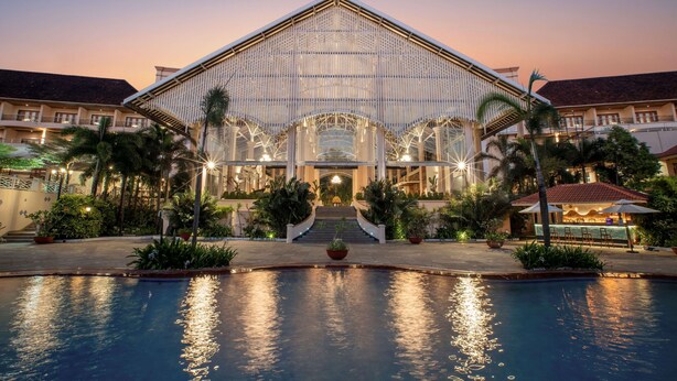 Um hotel com uma piscina grande, uma escadaria central e uma entrada coberta com guarda-corpos de vidro.