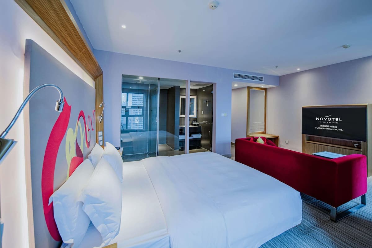 Quarto de hotel com uma cama, um sofá vermelho, uma TV e uma porta de vidro que dá para um banheiro.