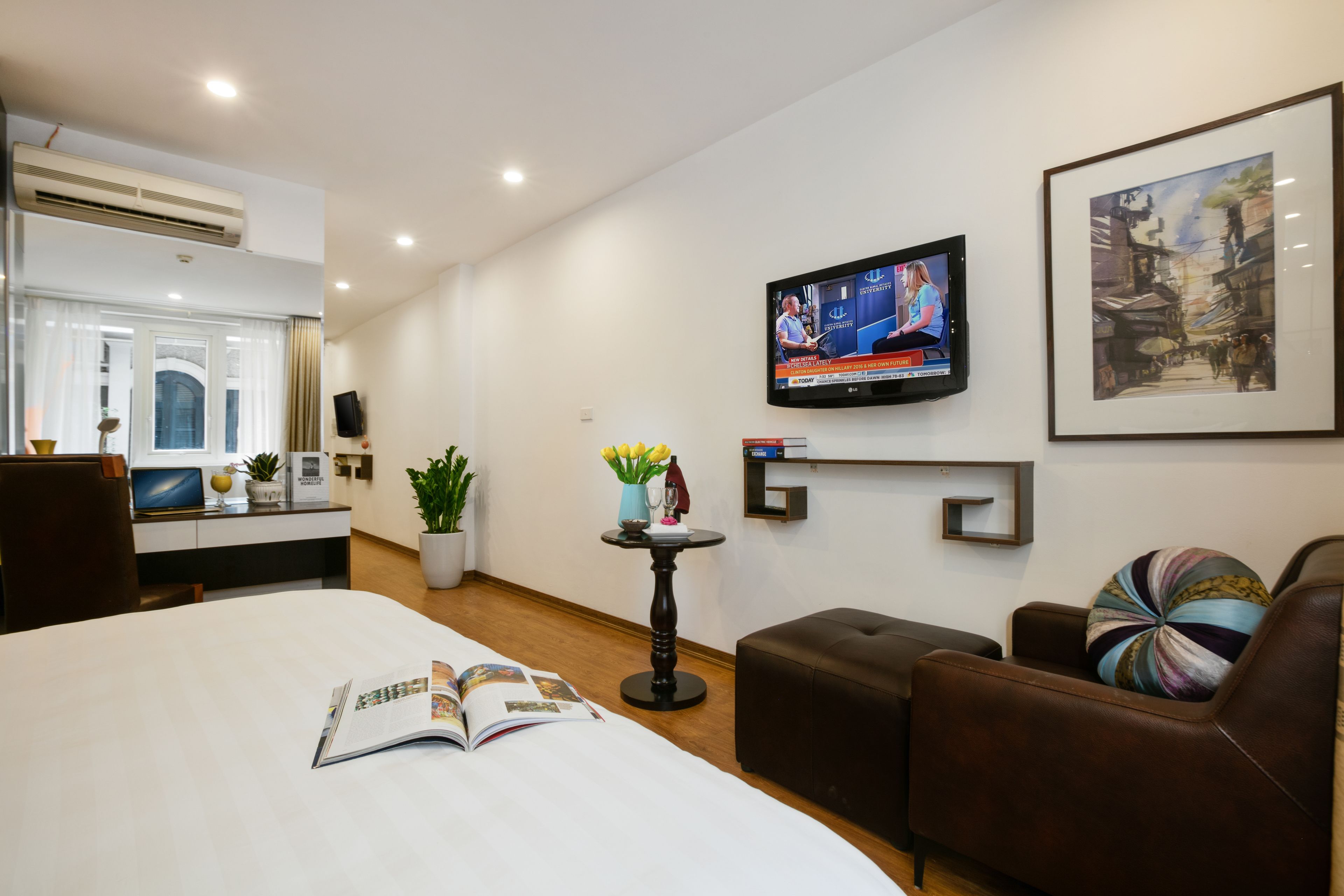Quarto de hotel moderno com uma cama grande, uma TV de tela plana na parede, uma mesa pequena com um vaso de flores e um quadro emoldurado na parede.