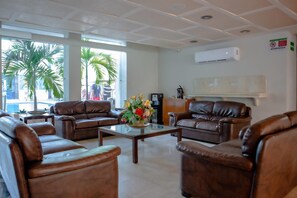 Um saguão de hotel com sofás de couro, uma mesa de centro de vidro e uma janela grande com palmeiras lá fora.