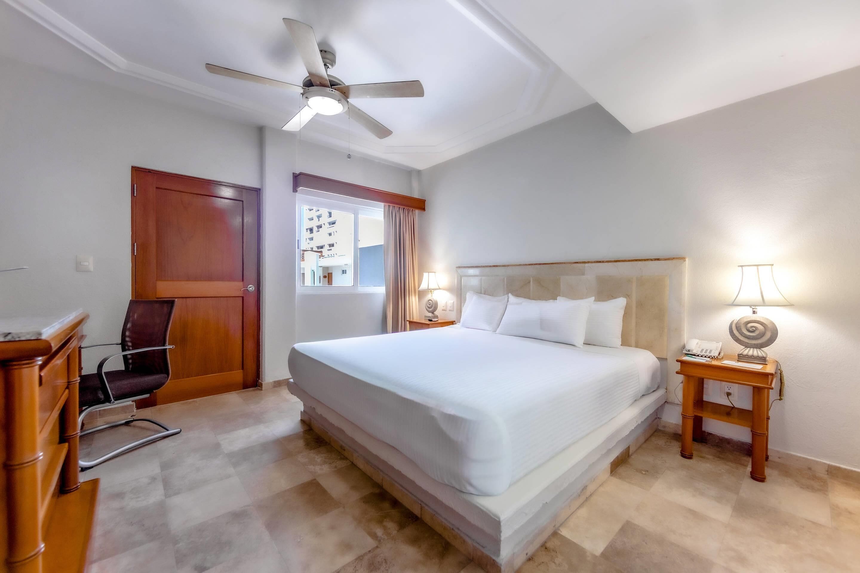 Quarto com cama, escrivaninha, cadeira, ventoinha no teto, janela e mesa de cabeceira com abajur.