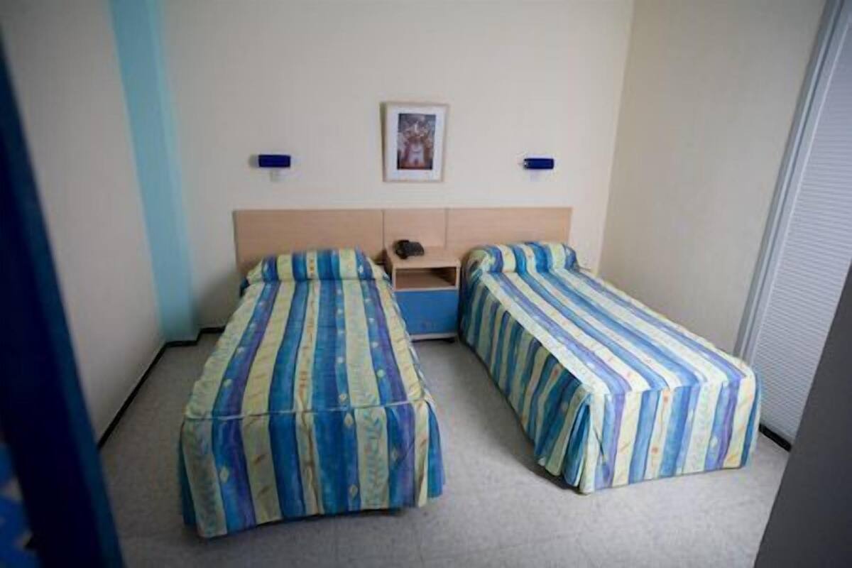 Quarto de hotel com duas camas de solteiro, uma escrivaninha pequena e um quadro na parede.