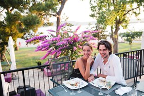 Um casal jantando ao ar livre, com uma mesa posta para dois, cercado por vegetação exuberante e flores roxas.