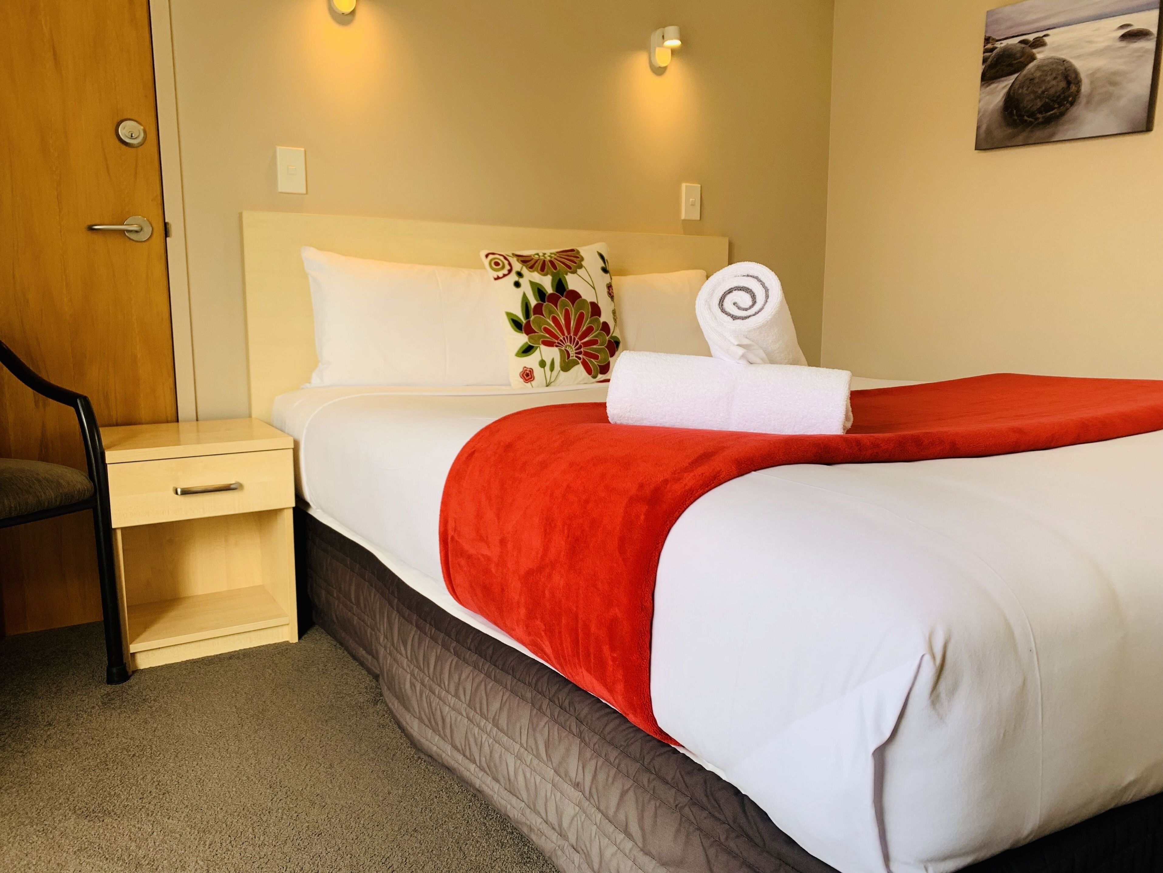 Quarto de hotel com cama, mesa de cabeceira, uma cadeira e luminária na parede.