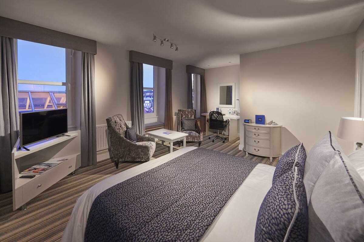 Quarto de hotel com uma cama grande, televisão, escrivaninha, duas cadeiras, cômoda e janela com vista para a cidade.