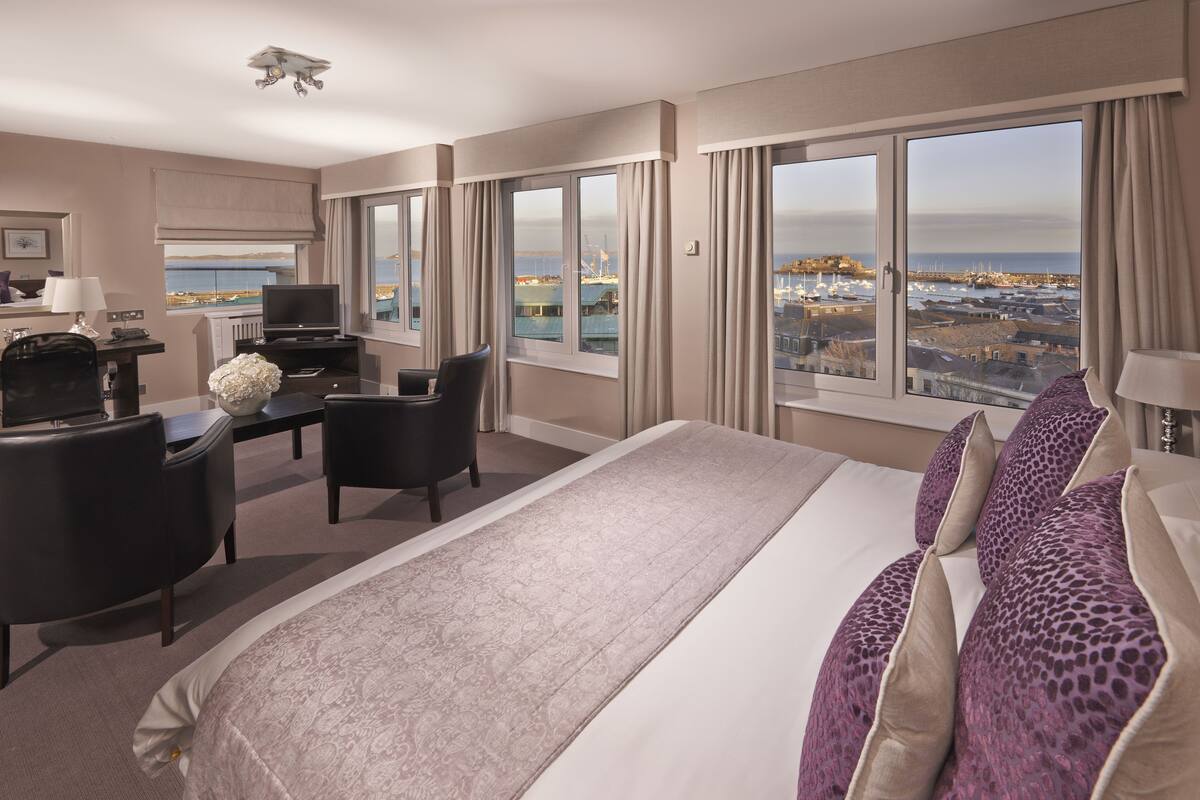 Quarto de hotel com uma cama grande, uma mesa com televisão, uma cadeira e vista para a cidade e o mar.