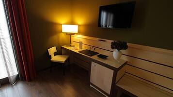 Quarto de hotel compacto com uma escrivaninha de madeira, uma cadeira, um armário pequeno, uma mesa de cabeceira com um vaso e uma televisão de tela plana fixada na parede.