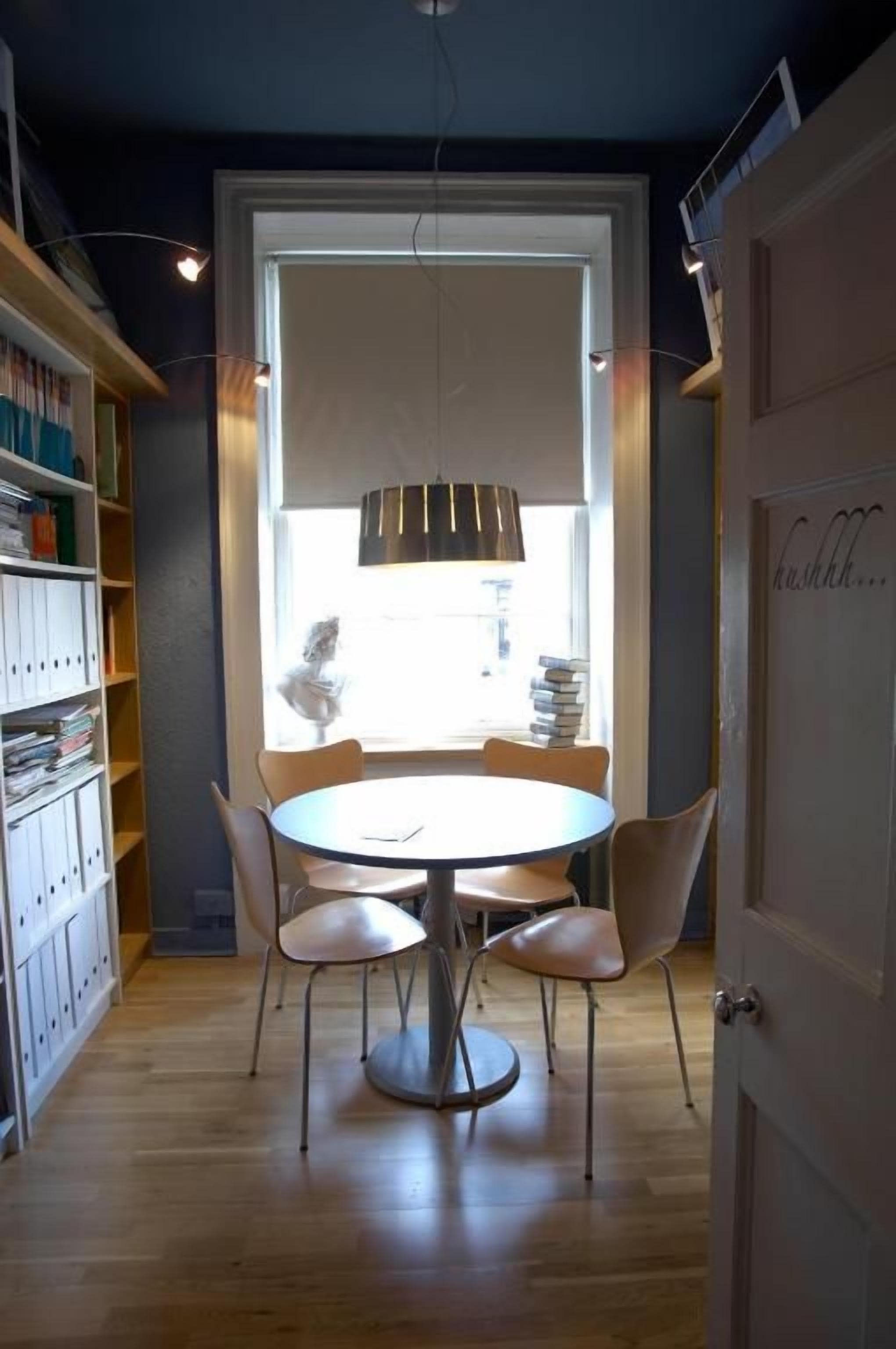 Um quarto com uma mesa redonda e quatro cadeiras, uma estante de livros e uma porta com um letreiro.