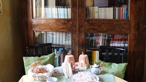 Uma mesa de jantar posta com serviço de chá, cercada por cadeiras e uma estante de madeira com livros.