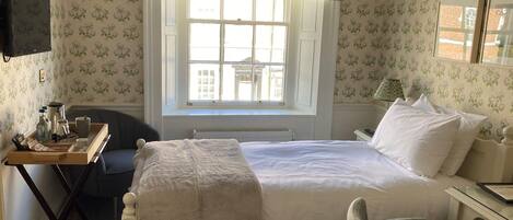 Quarto de hotel com cama, escrivaninha com cadeira, janela com cortinas e radiador.