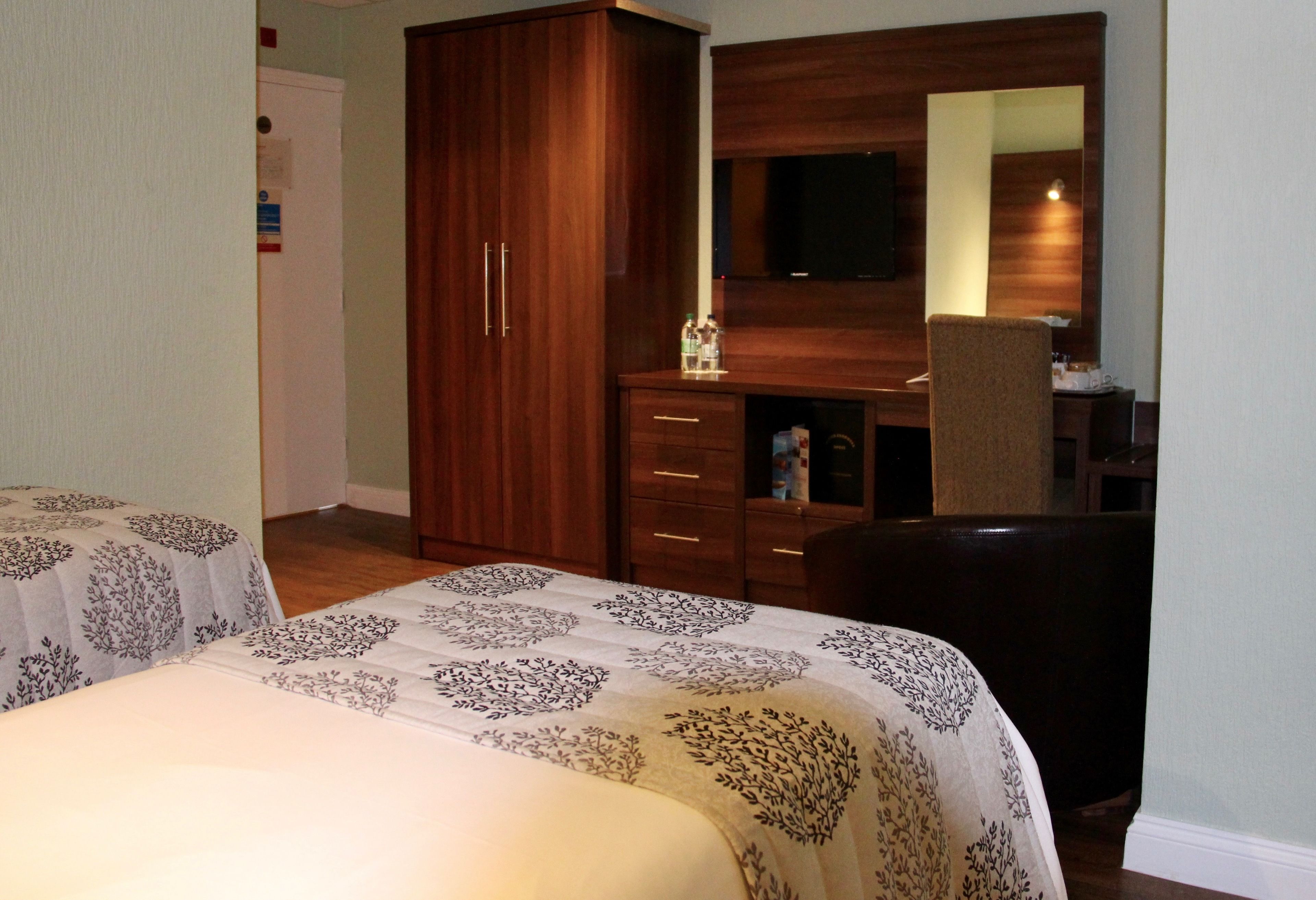 Quarto de hotel com uma cama, uma escrivaninha com cadeira, um guarda-roupa e uma televisão.