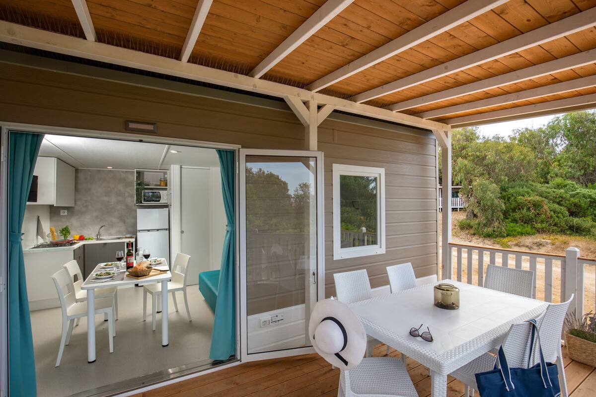 Uma área de refeições ao ar livre coberta, com mesa e cadeiras, uma porta de correr de vidro que dá para uma cozinha, e vista para o verde ao redor.