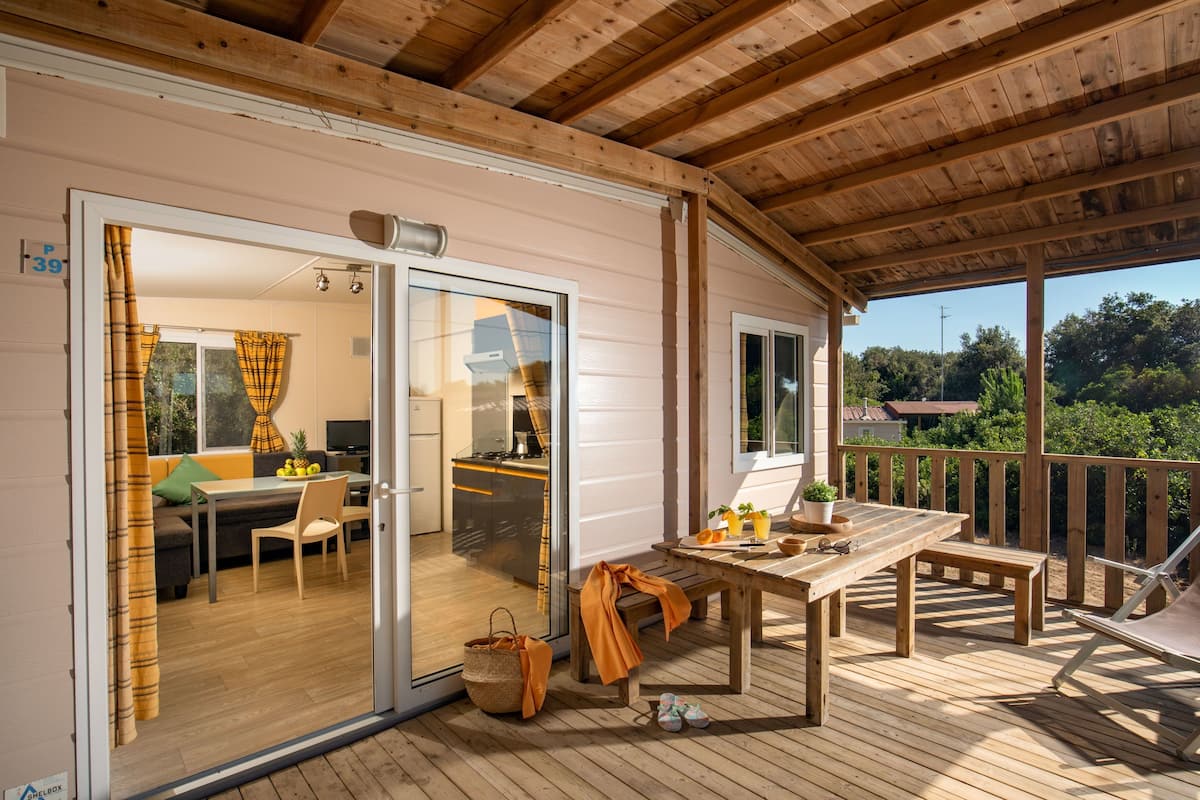 Um deck de madeira com mesa e cadeiras, uma porta de vidro deslizante que dá para uma cozinha, e vista para vegetação.