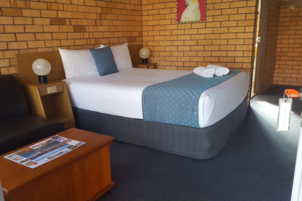 Quarto de hotel com cama, mesa de cabeceira de madeira com abajur, pequena mesa com um folheto e parede de tijolos.