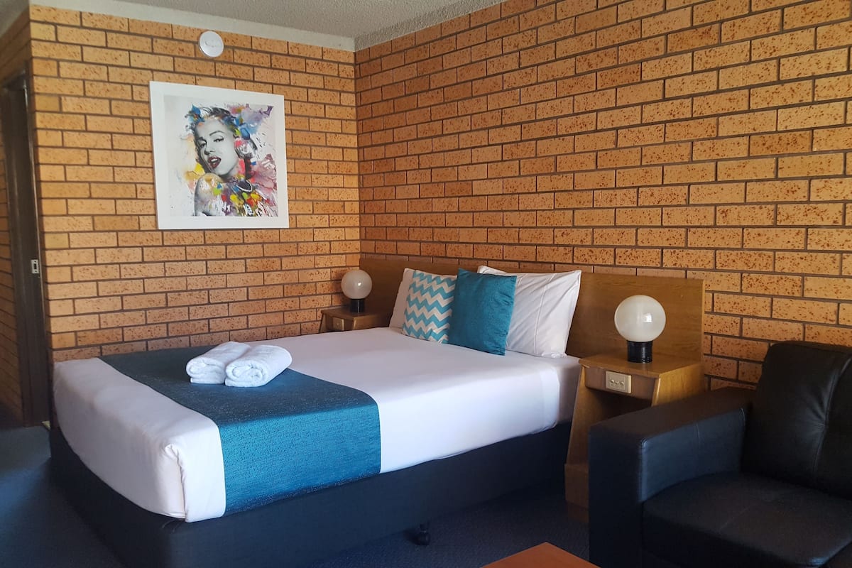 Quarto de hotel com parede em padrão tijolo, cama com roupa de cama branca, travesseiro azul de destaque, poltrona de couro preta e mesa de cabeceira com luminária.
