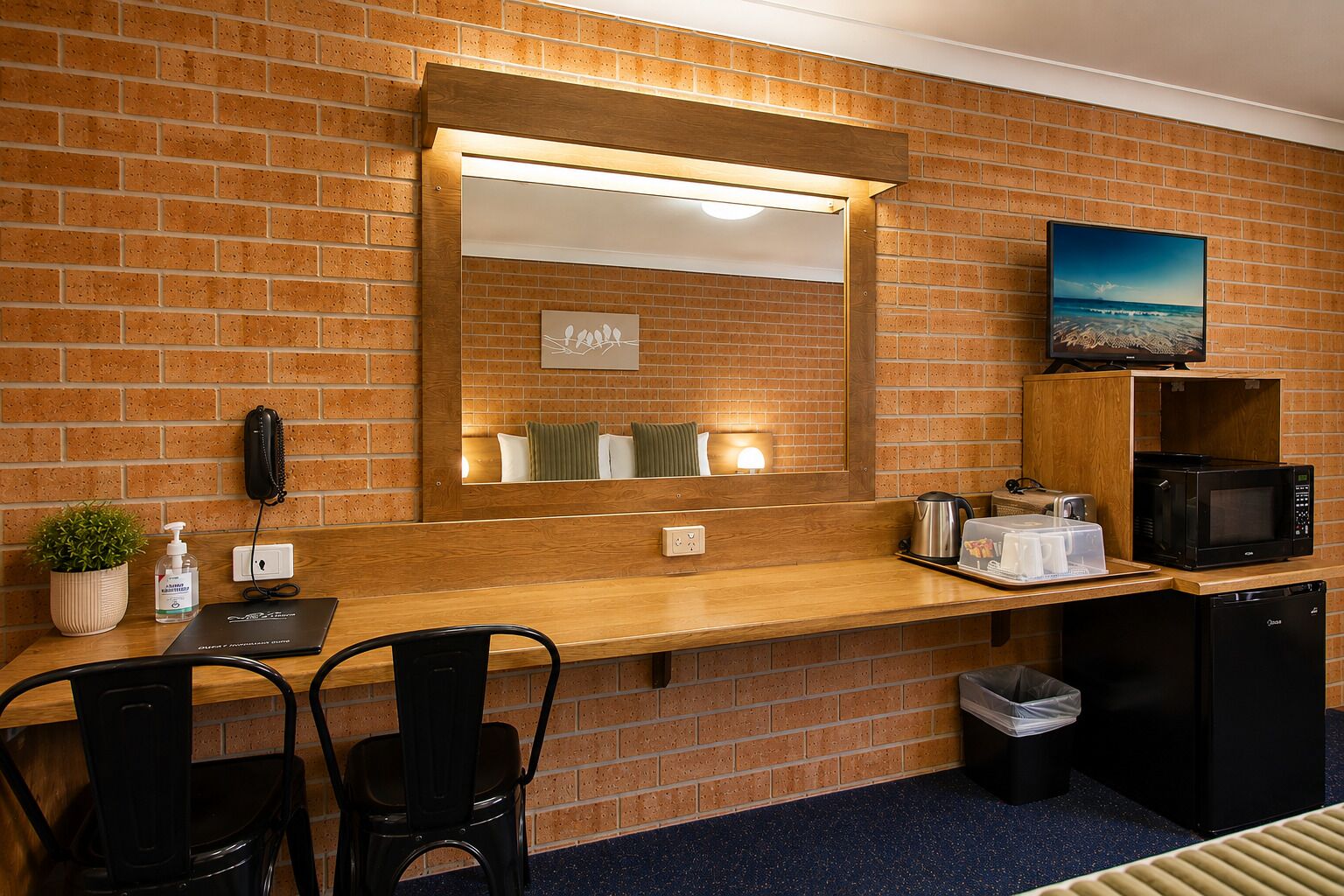 Quarto de hotel com escrivaninha de madeira, espelho, micro-ondas, lixeira e uma pequena planta.