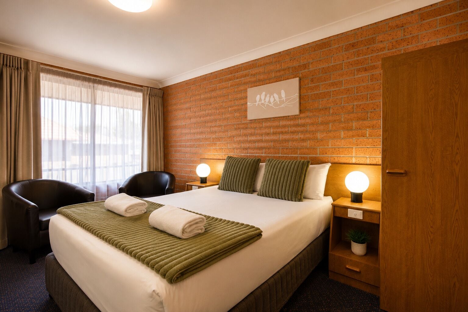 Quarto de hotel com parede de tijolos, cama com roupa de cama verde e branca, duas luminárias de cabeceira, janela com cortinas e uma cadeira.
