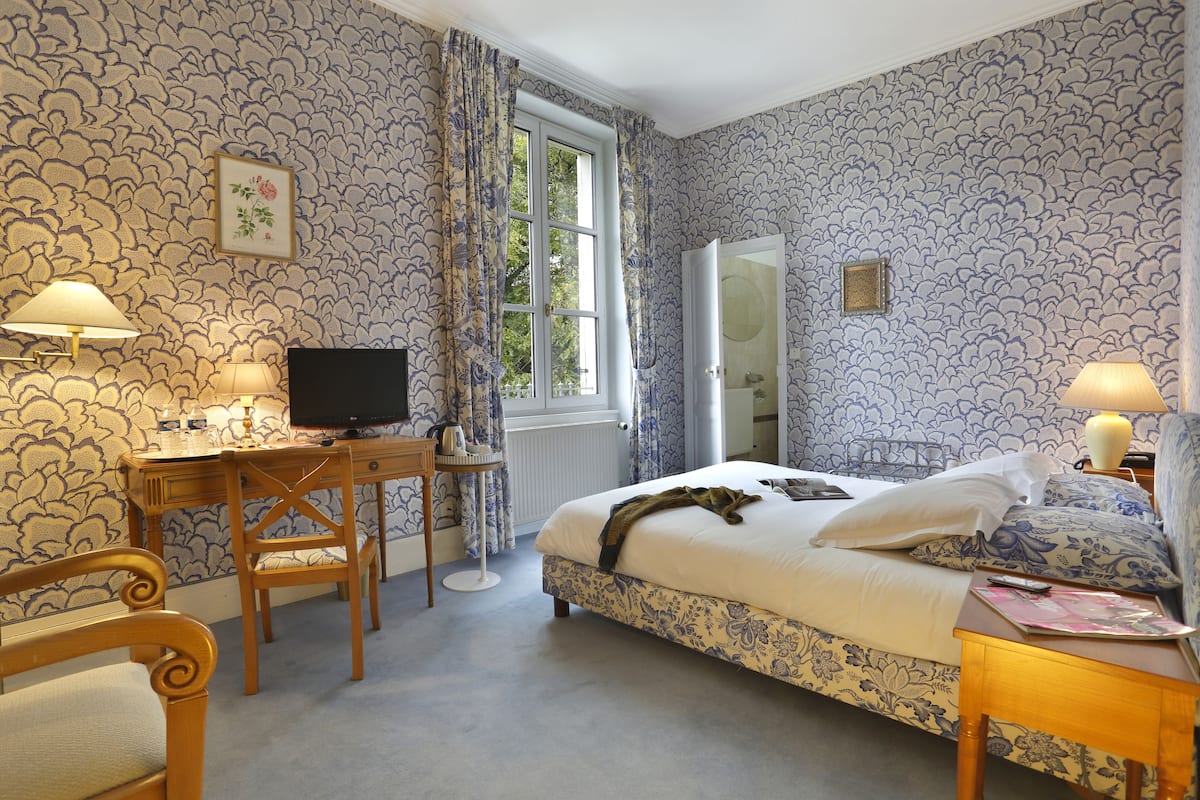 Quarto com cama, escrivaninha, cadeira, televisão e janela com cortinas.